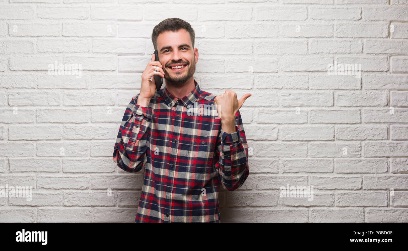 Young adult man talking on the phone standing over white brick wall ...