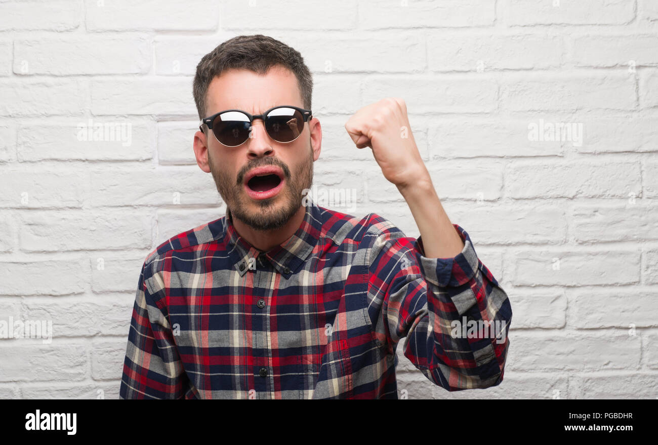 Young adult man wearing sunglasses standing over white brick wall ...