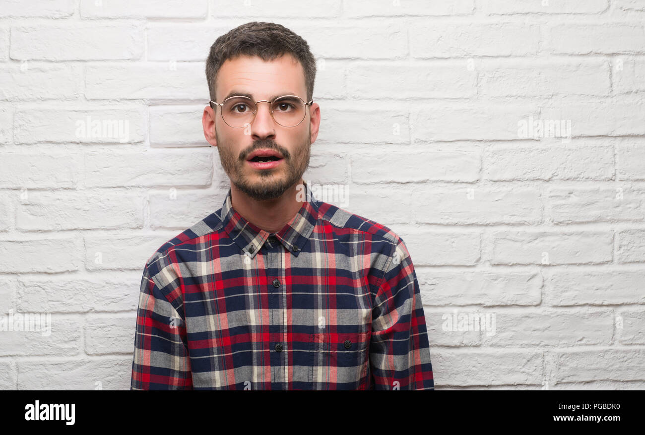 Young adult man standing over white brick wall scared in shock with a ...