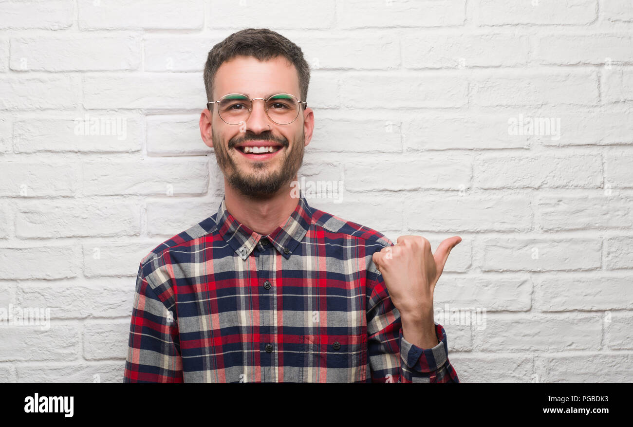 Young adult man standing over white brick wall pointing and showing ...