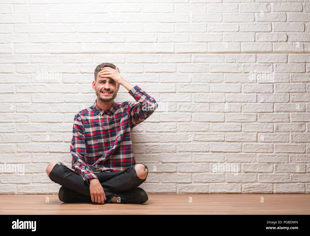 Young adult man sitting over white brick wall stressed with hand on ...
