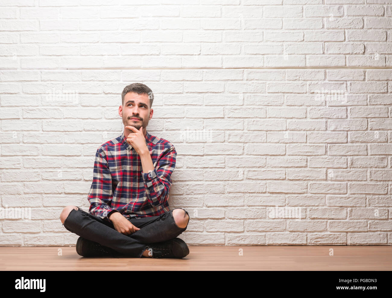 Young adult man sitting over white brick wall serious face thinking ...