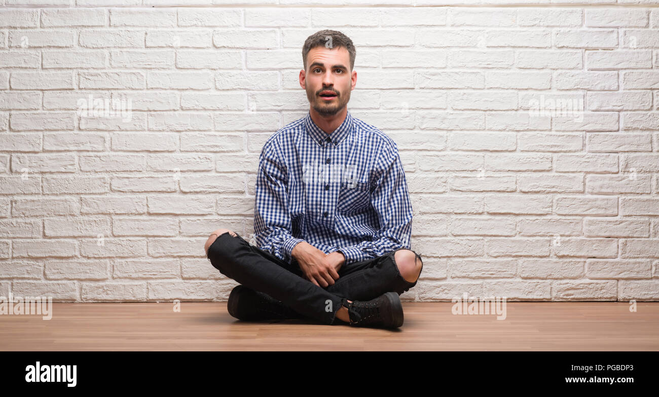 Young adult man sitting over white brick wall afraid and shocked with ...