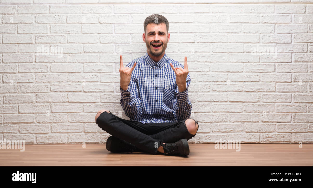 Young adult man sitting over white brick wall shouting with crazy ...