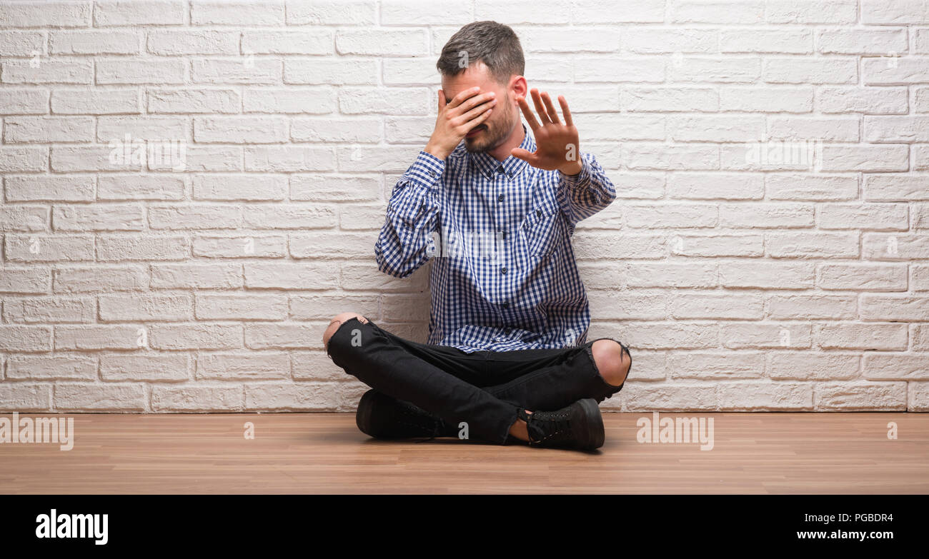 Young adult man sitting over white brick wall covering eyes with hands ...