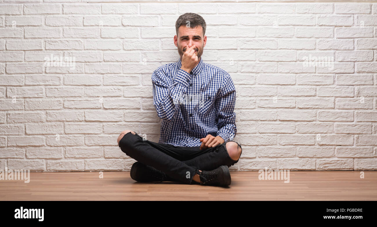 Young adult man sitting over white brick wall smelling something stinky ...