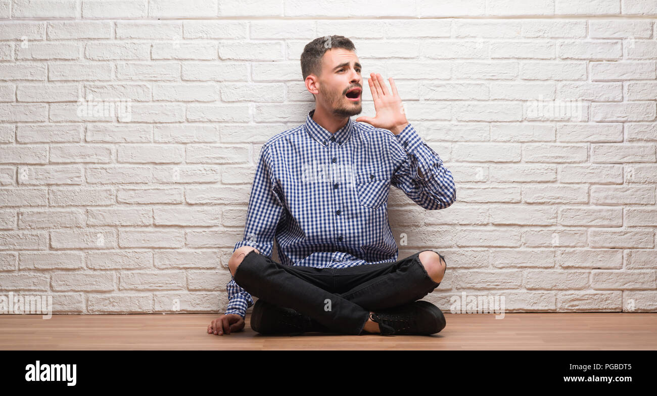 Young adult man sitting over white brick wall shouting and screaming ...