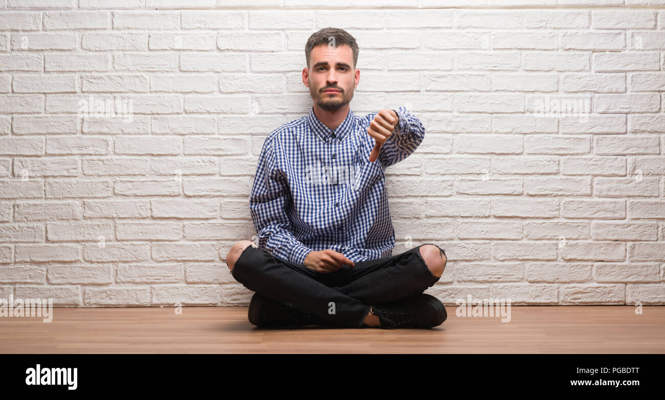 Young adult man sitting over white brick wall looking unhappy and angry ...