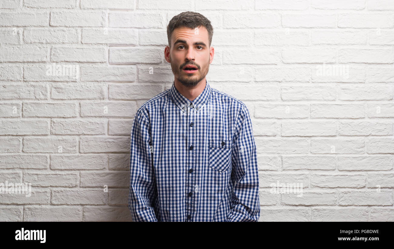Young adult man standing over white brick wall scared in shock with a ...