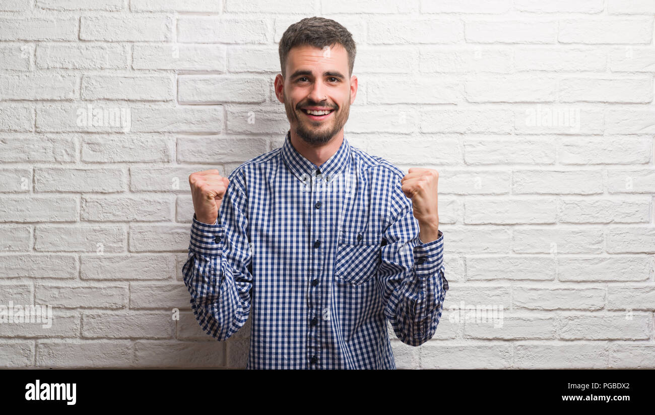 Young adult man standing over white brick wall screaming proud and ...