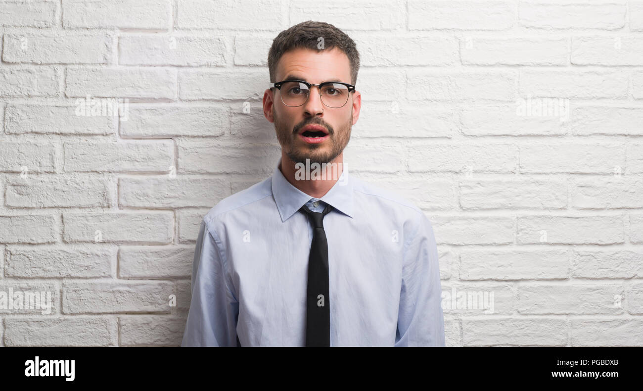 Young adult business man standing over white brick wall scared in shock ...