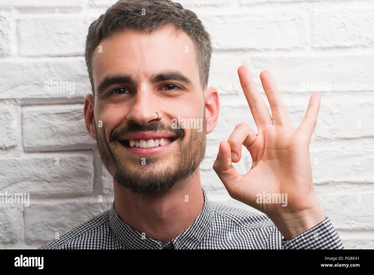 Young adult man standing over white brick wall doing ok sign with ...