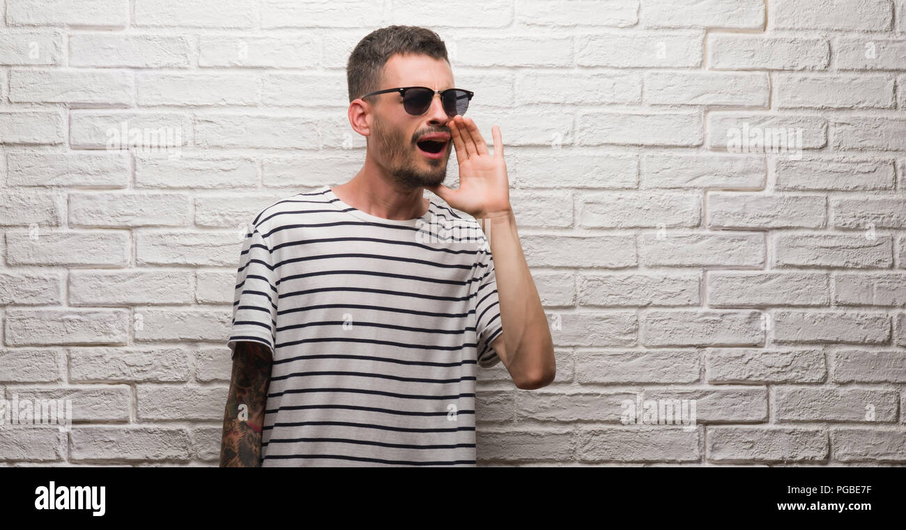 Young adult man wearing sunglasses standing over white brick wall ...