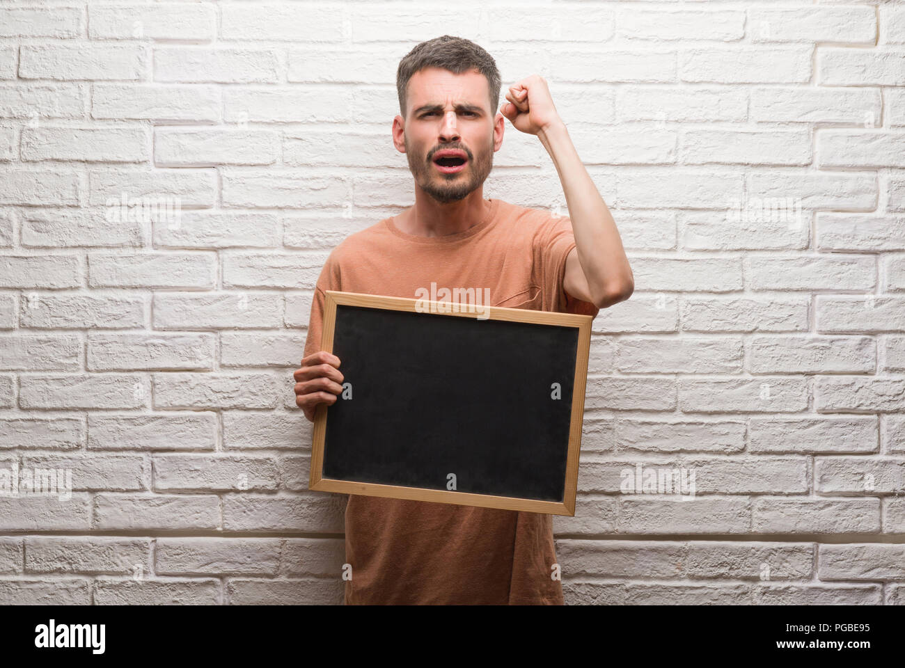 Young adult man over brick wall holding blackboard annoyed and ...