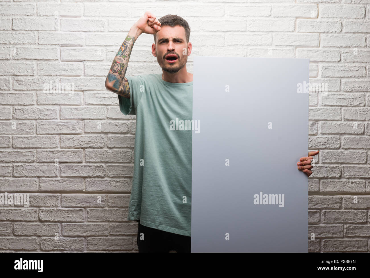 Young adult man over brick wall holding banner annoyed and frustrated ...
