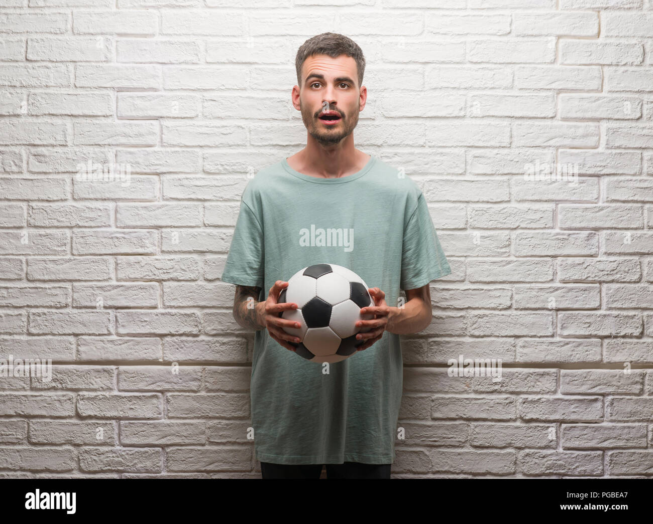Young adult man holding soccer ball standing over white brick wall ...