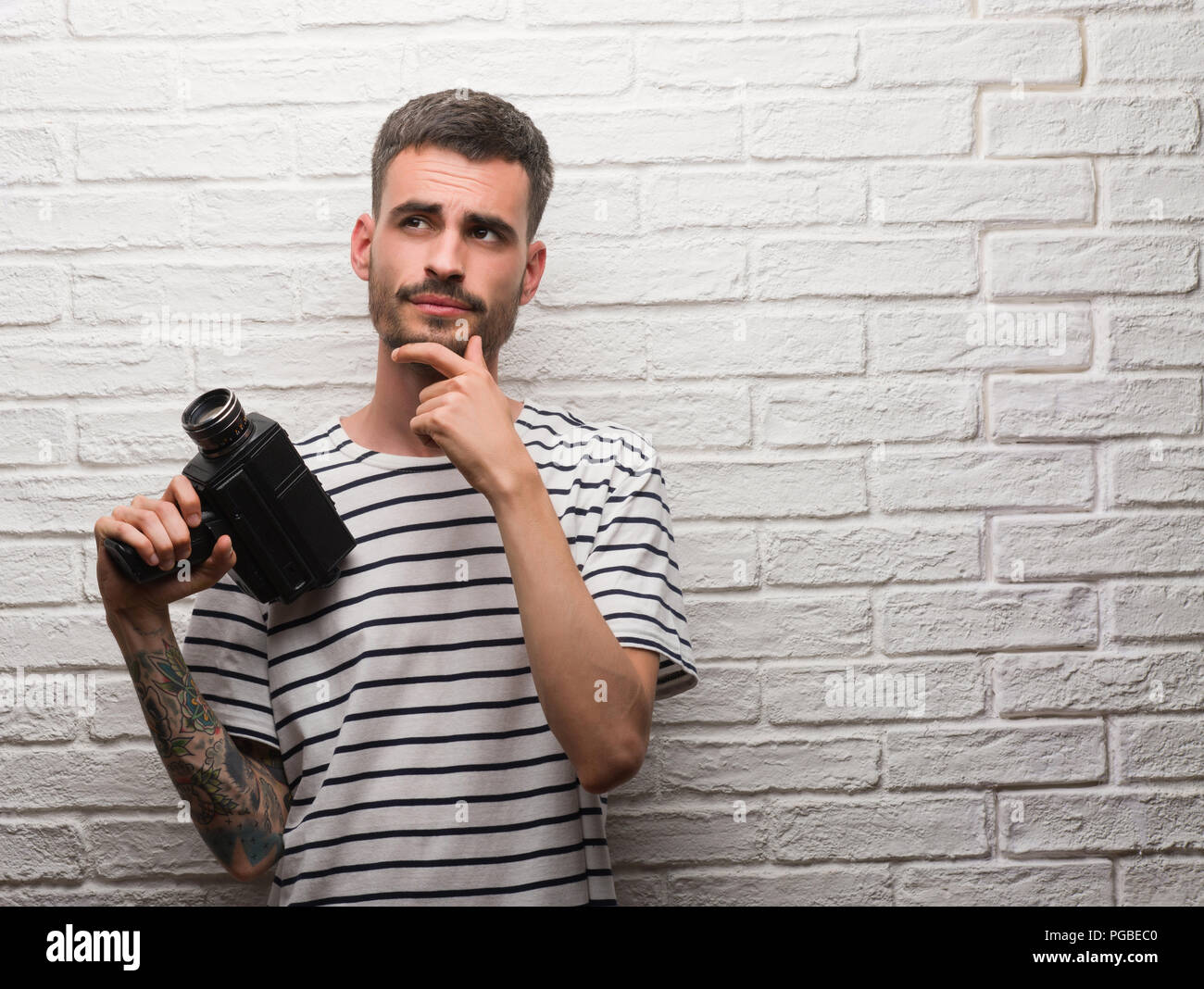 Young man filming holding video camera standing over white brick wall ...