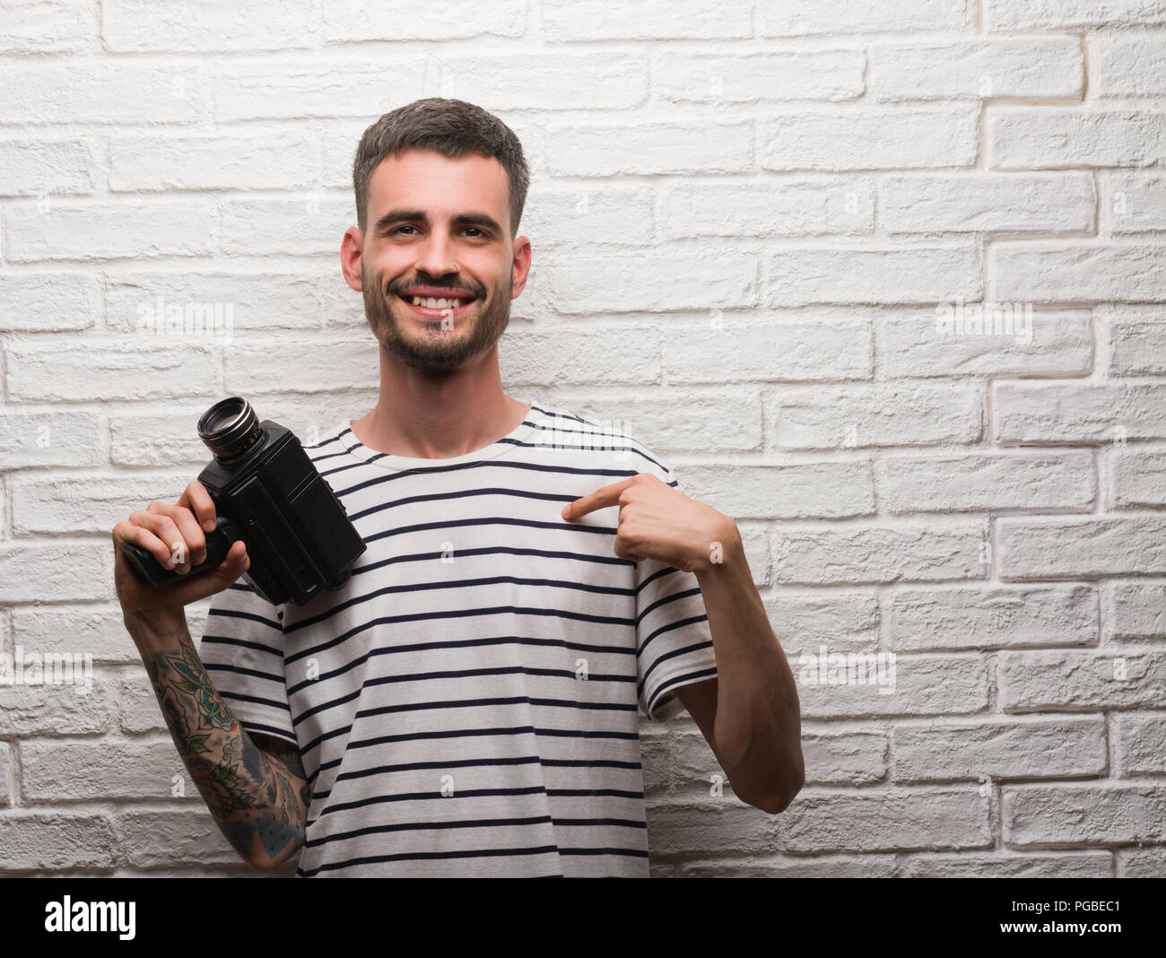 Young man filming holding video camera standing over white brick wall ...