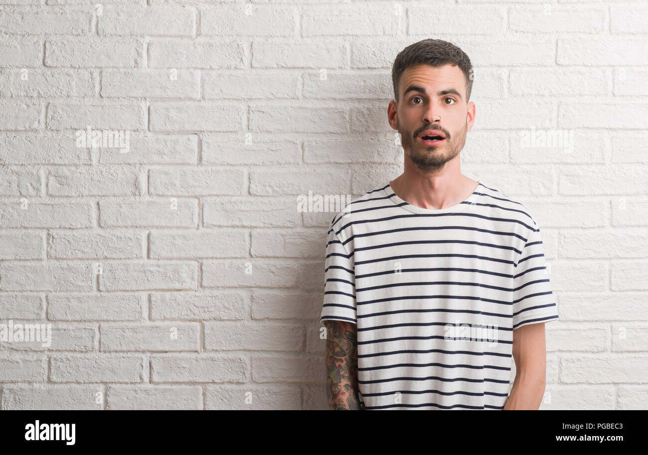 Young adult man standing over white brick wall scared in shock with a ...