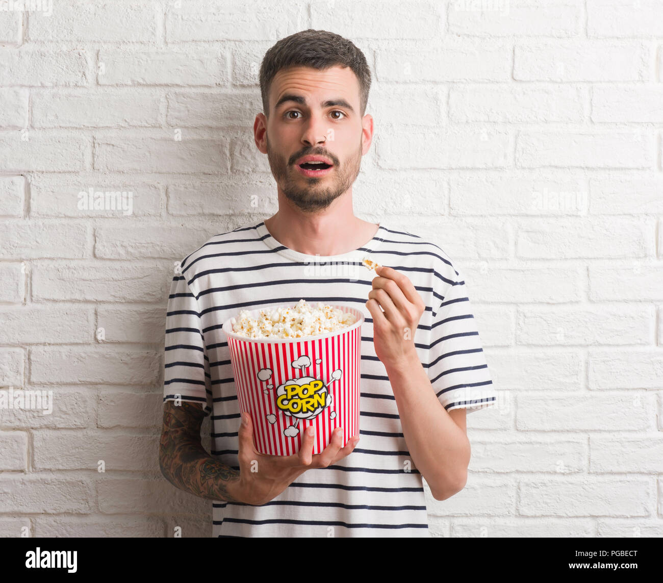 Young adult man eating popcorn standing over white brick wall scared in ...