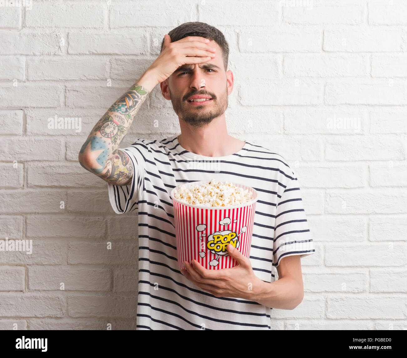 Young adult man eating popcorn standing over white brick wall stressed ...