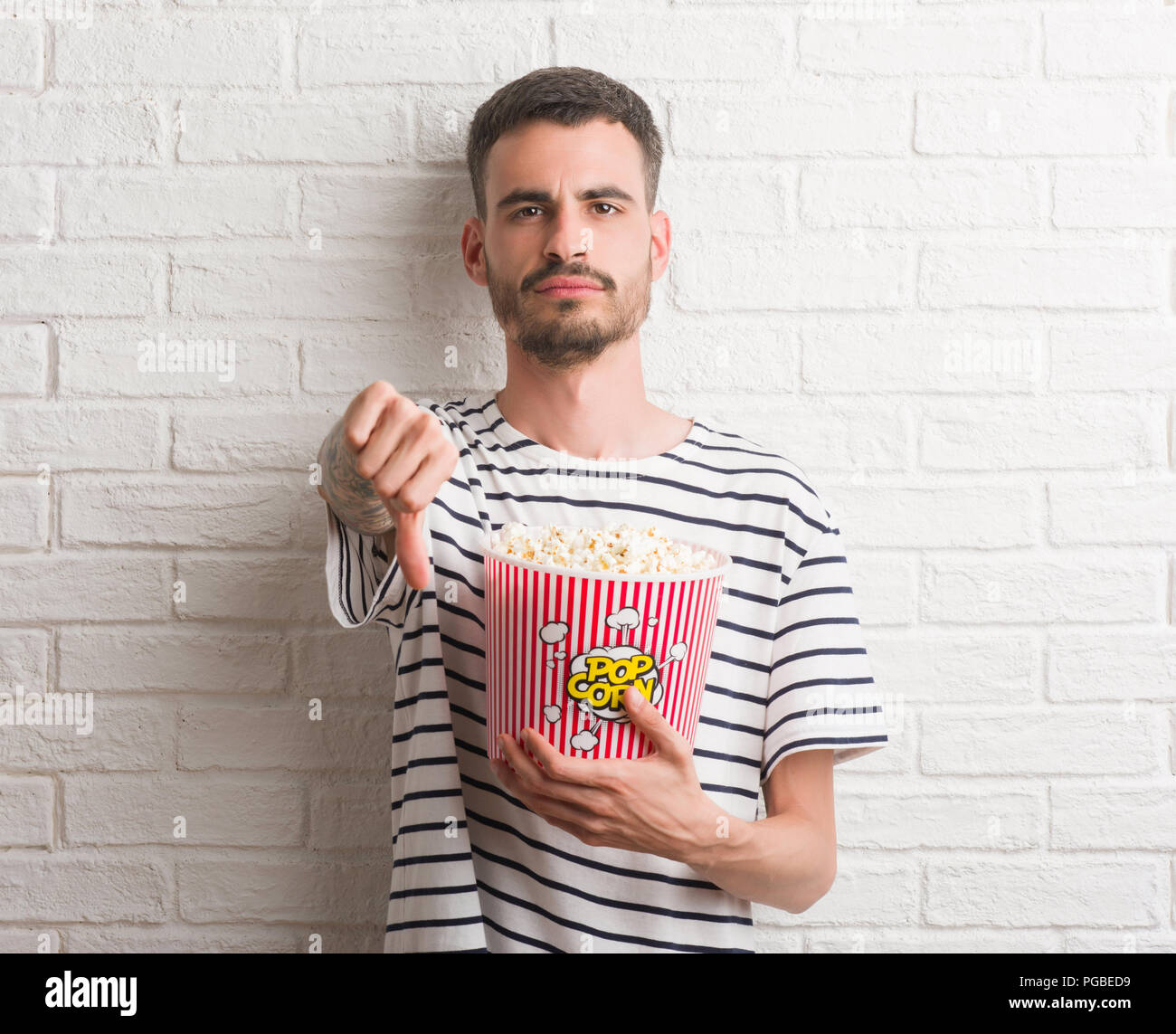 Young adult man eating popcorn standing over white brick wall with ...