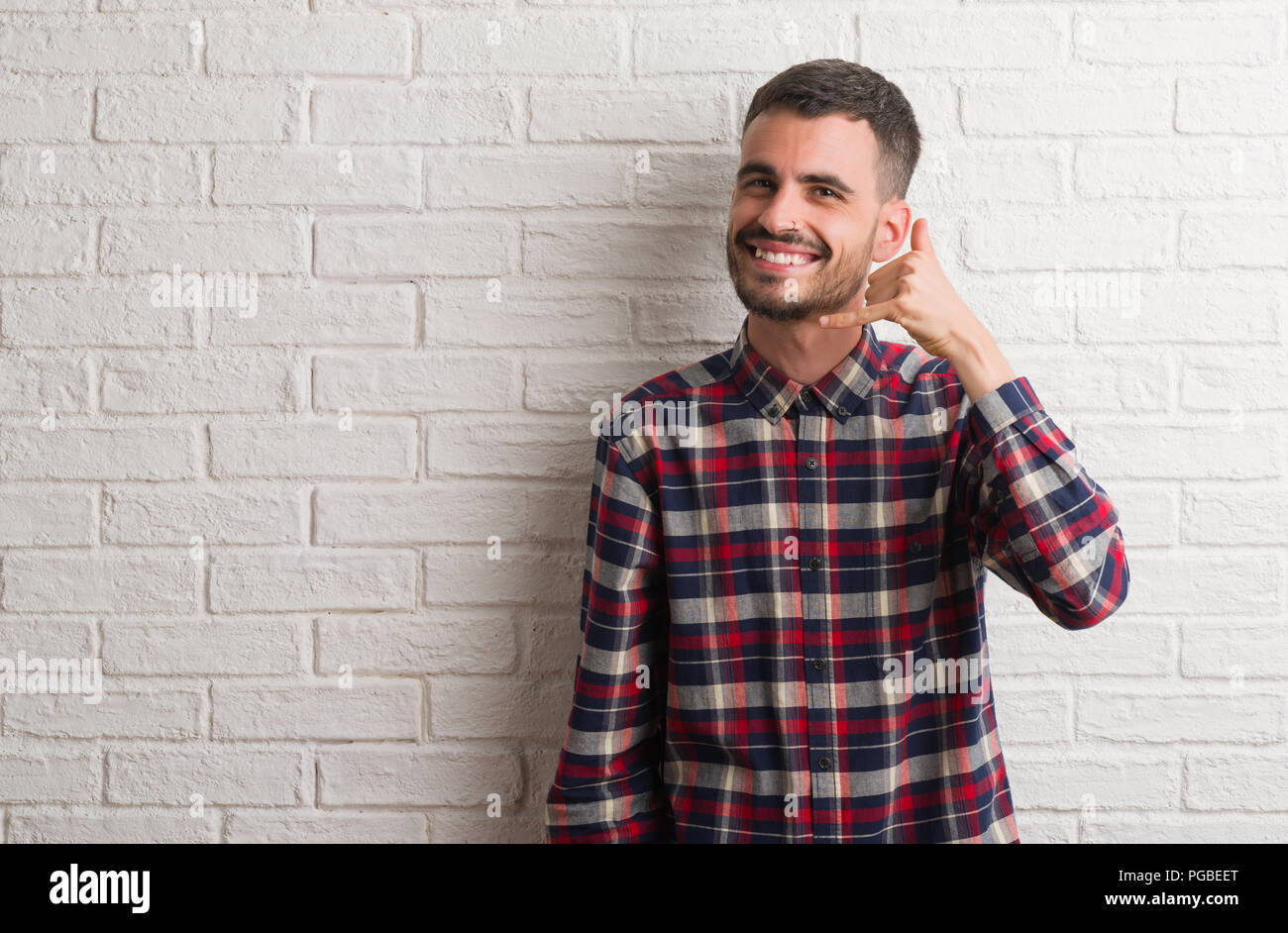 Young adult man standing over white brick wall smiling doing phone ...