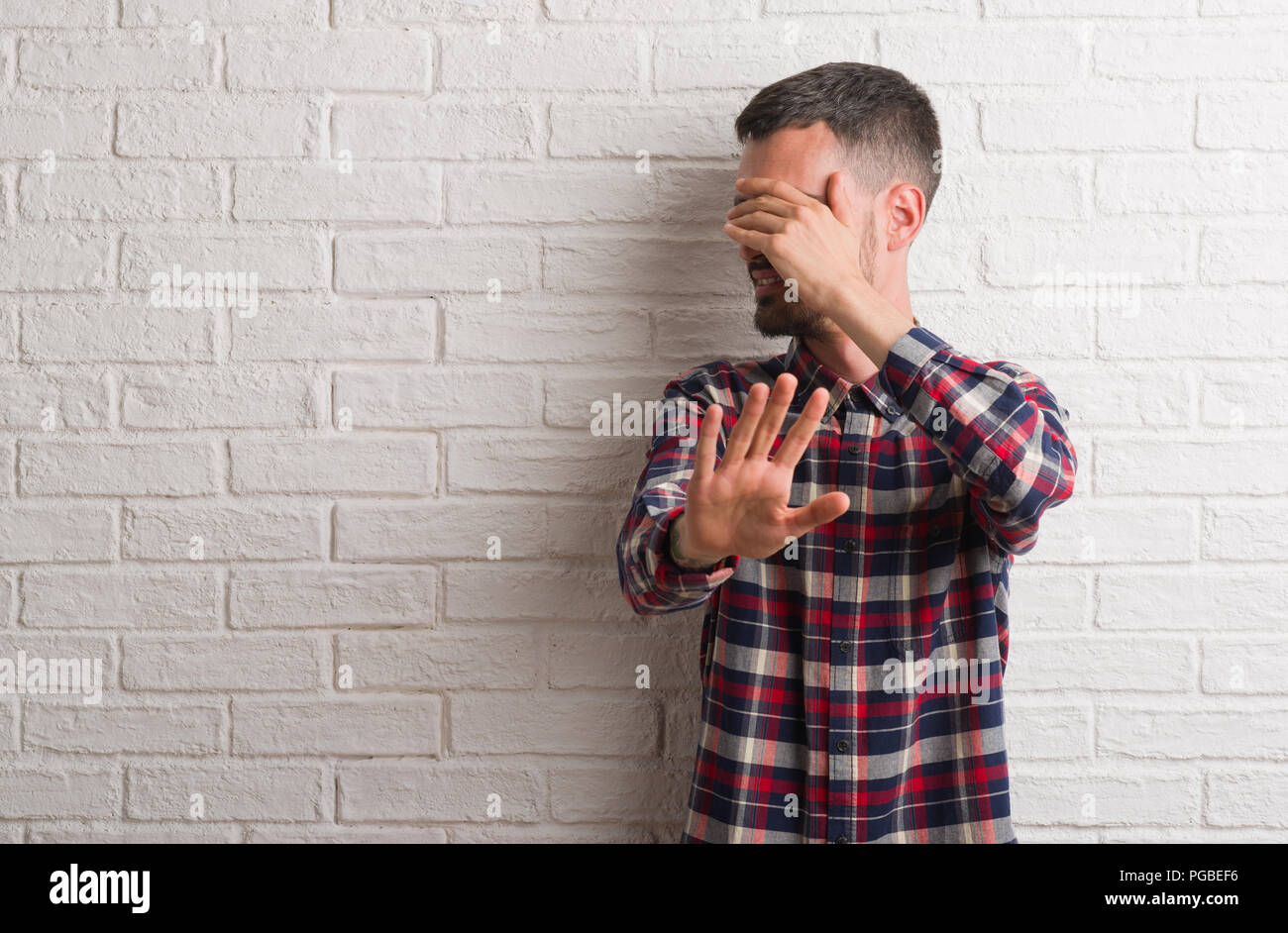 Young adult man standing over white brick wall covering eyes with hands ...