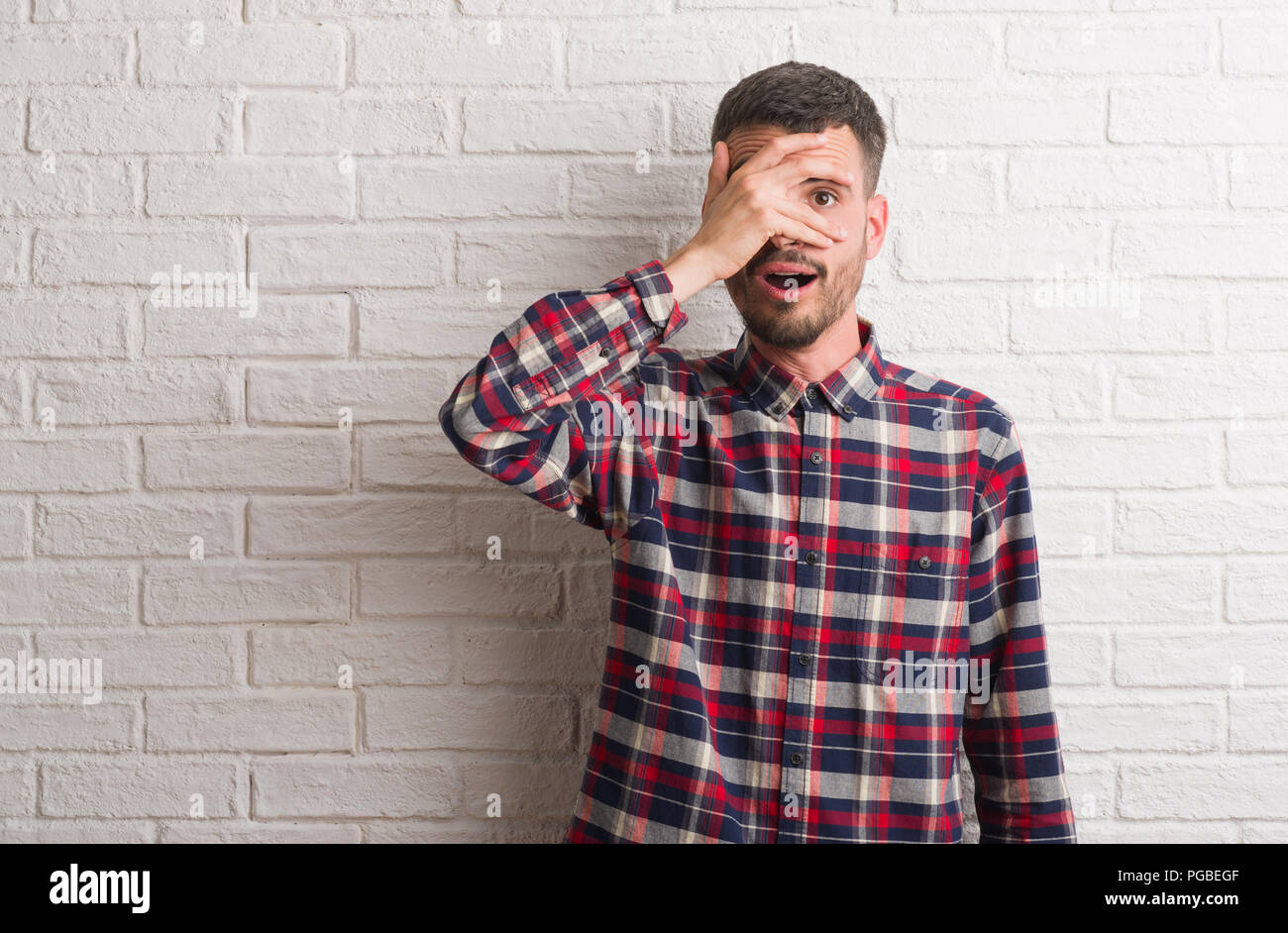 Young adult man standing over white brick wall peeking in shock ...