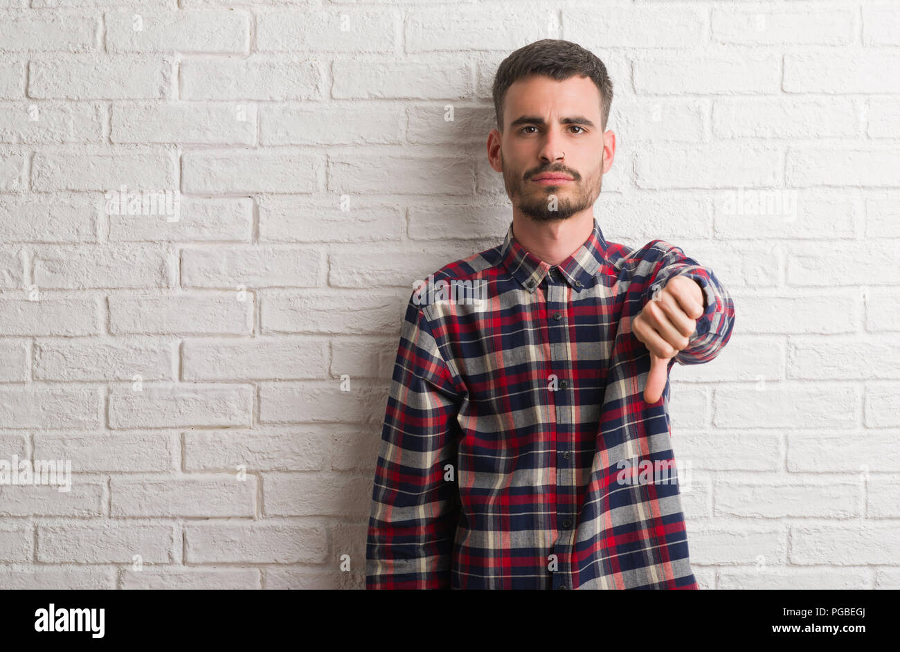 Young adult man standing over white brick wall looking unhappy and ...