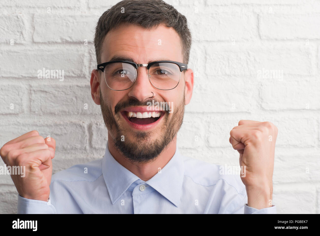 Young adult business man standing over white brick wall screaming proud ...