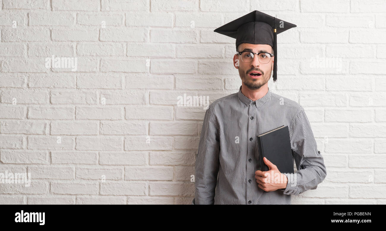 Young adult man over brick wall wearing graduation cap scared in shock ...