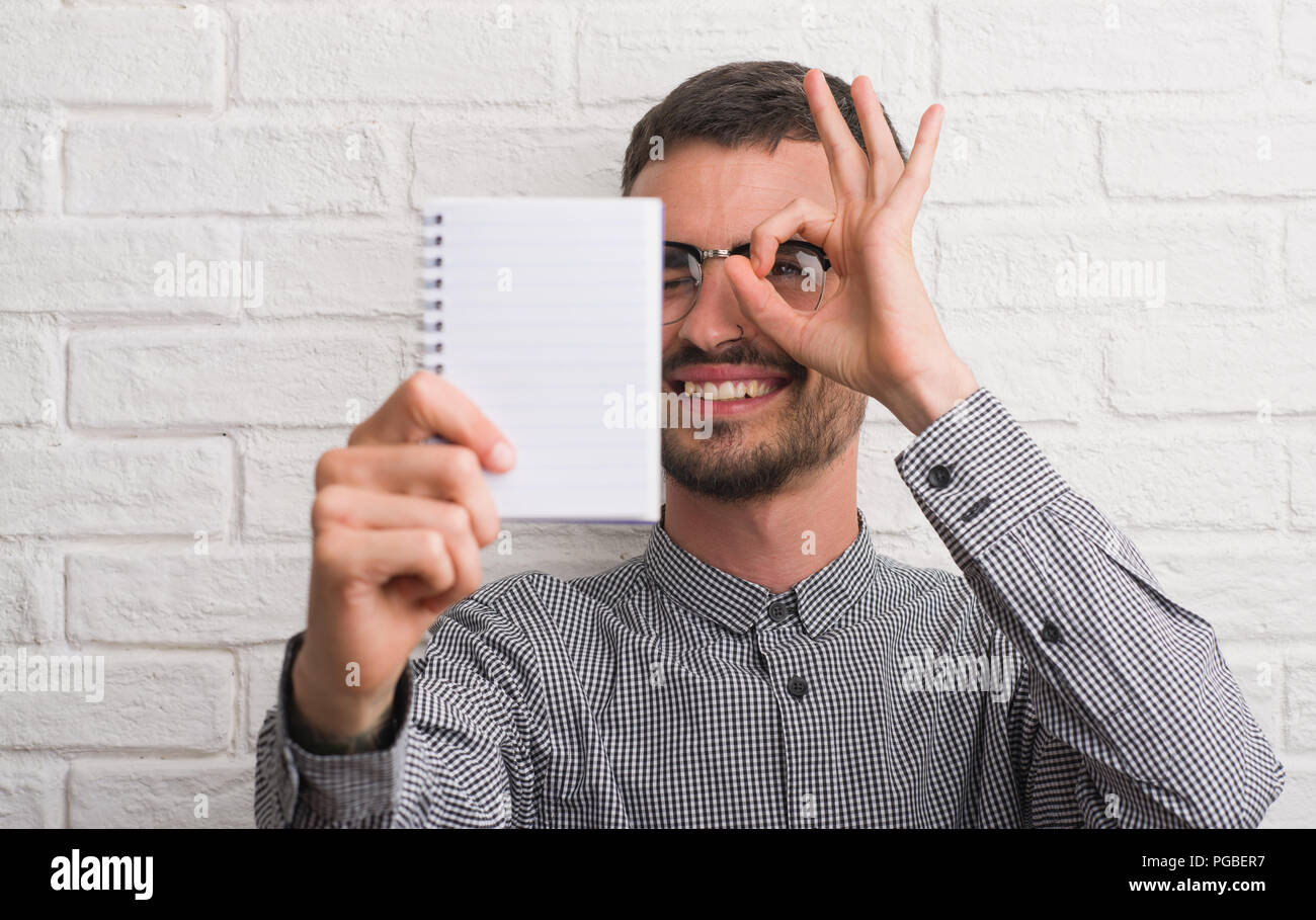 Young adult man holding notebook standing over white brick wall with ...