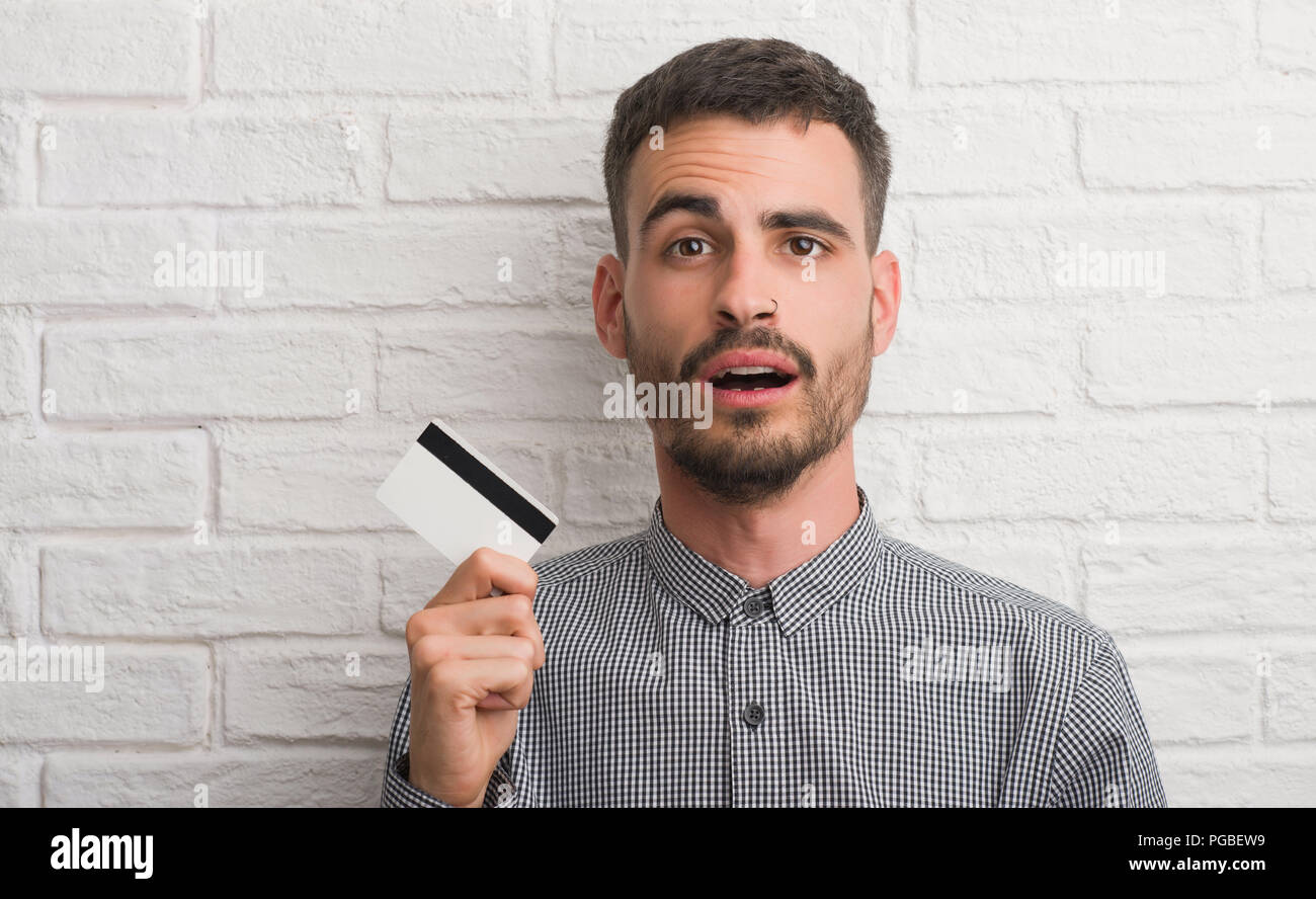 Young adult man over brick wall holding credit card scared in shock ...