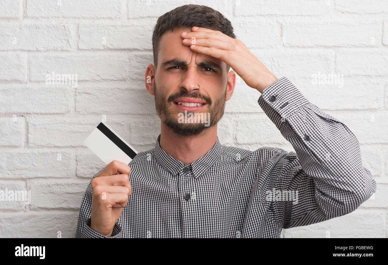 Young adult man over brick wall holding credit card stressed with hand ...