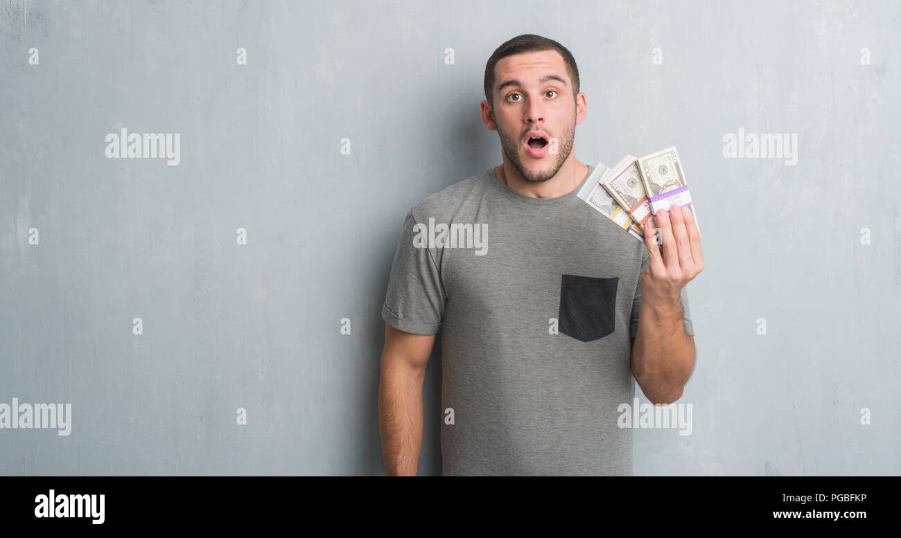 Young caucasian man over grey grunge wall holding a bunch of money ...
