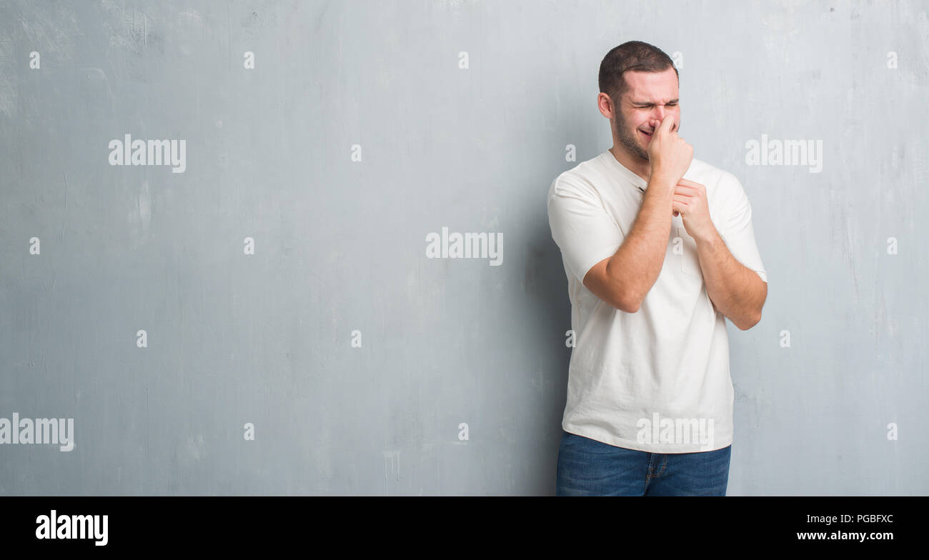 Young caucasian man over grey grunge wall wearing sunglasses smelling ...