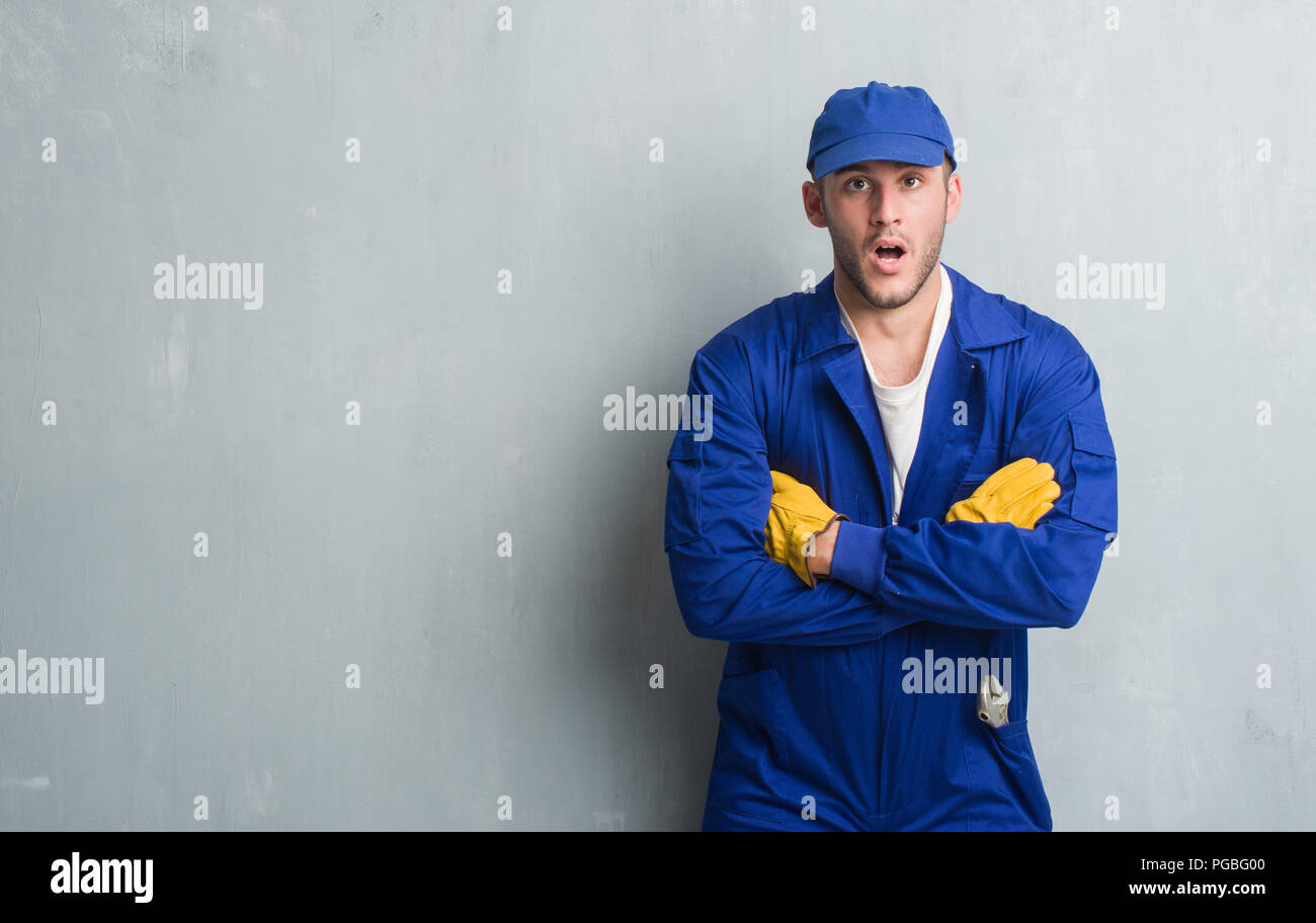 Young caucasian man over grey grunge wall wearing mechanic uniform ...
