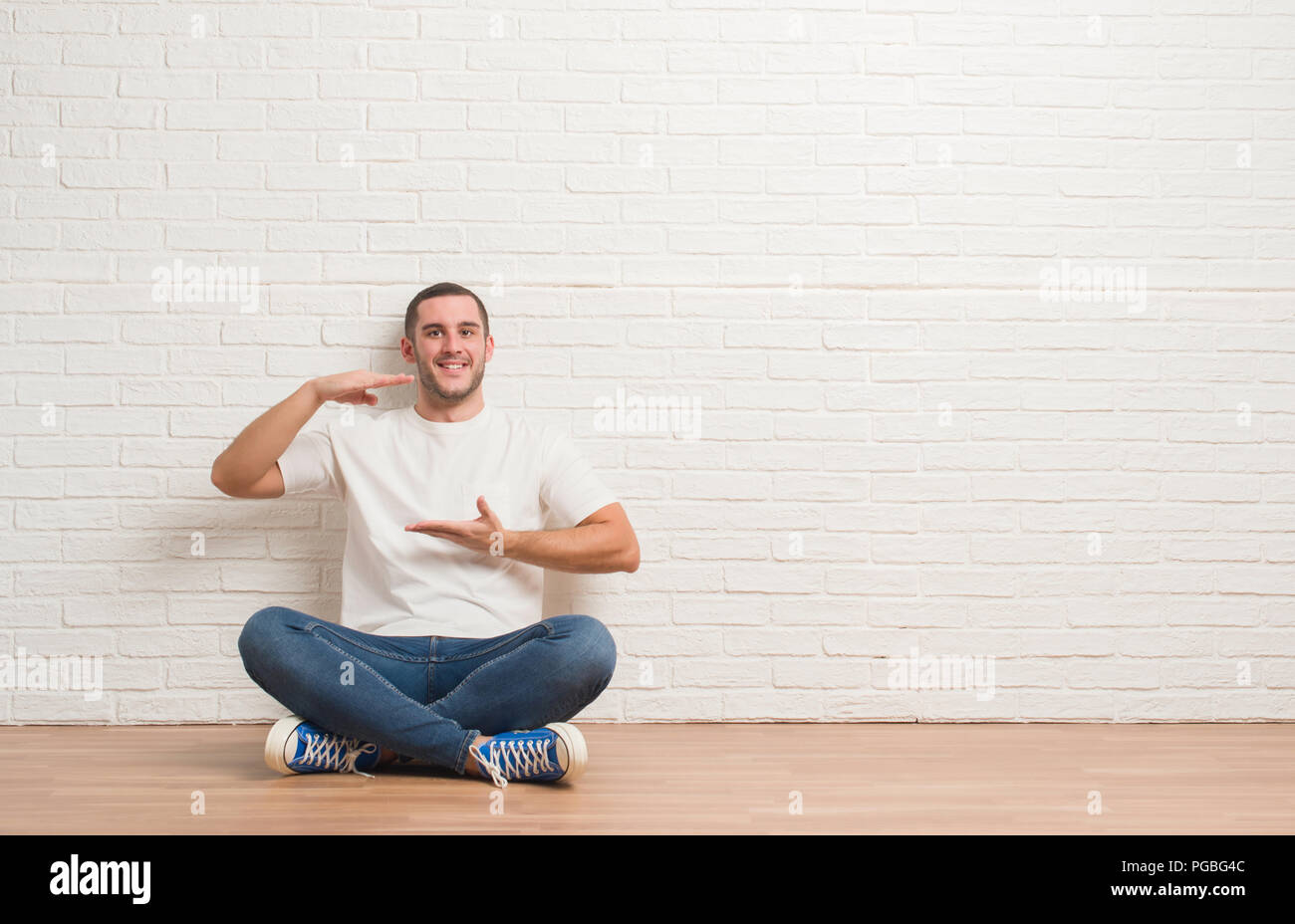 Young caucasian man sitting on the floor over white brick wall ...
