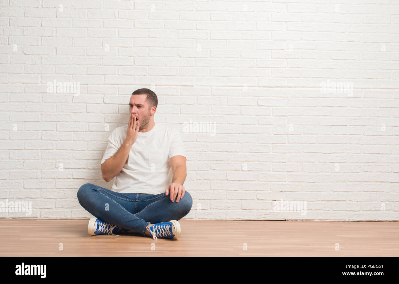 Young caucasian man sitting on the floor over white brick wall bored ...