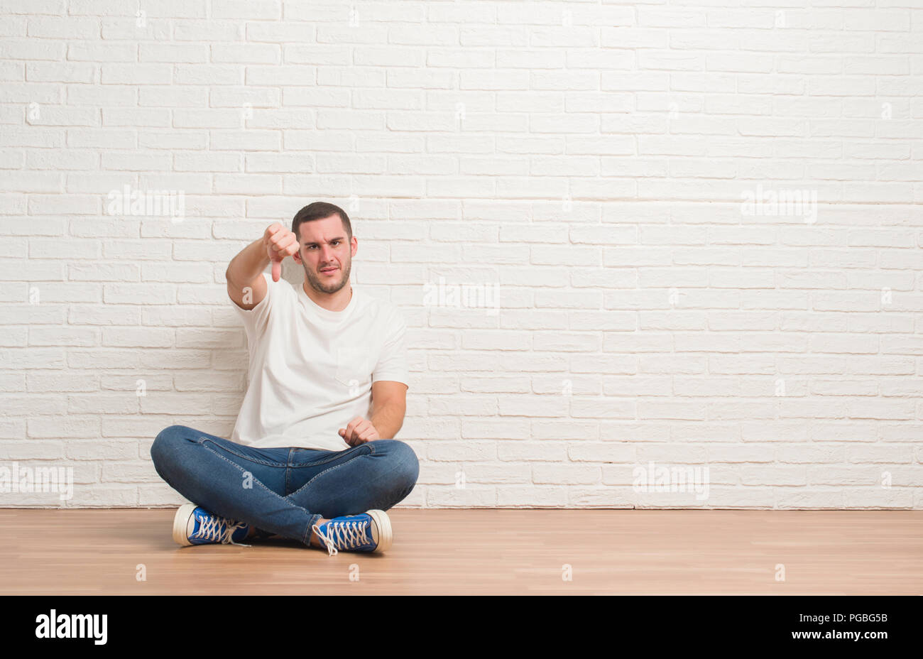 Young caucasian man sitting on the floor over white brick wall looking ...