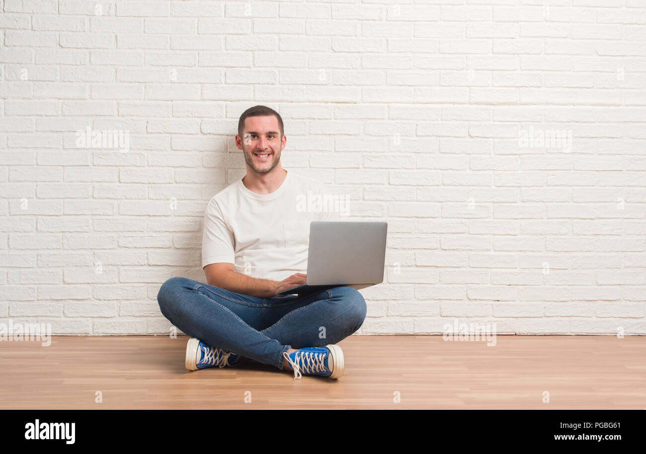 Young caucasian man sitting over white brick wall using computer laptop ...