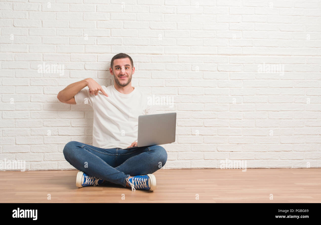 Young caucasian man sitting over white brick wall using computer laptop ...