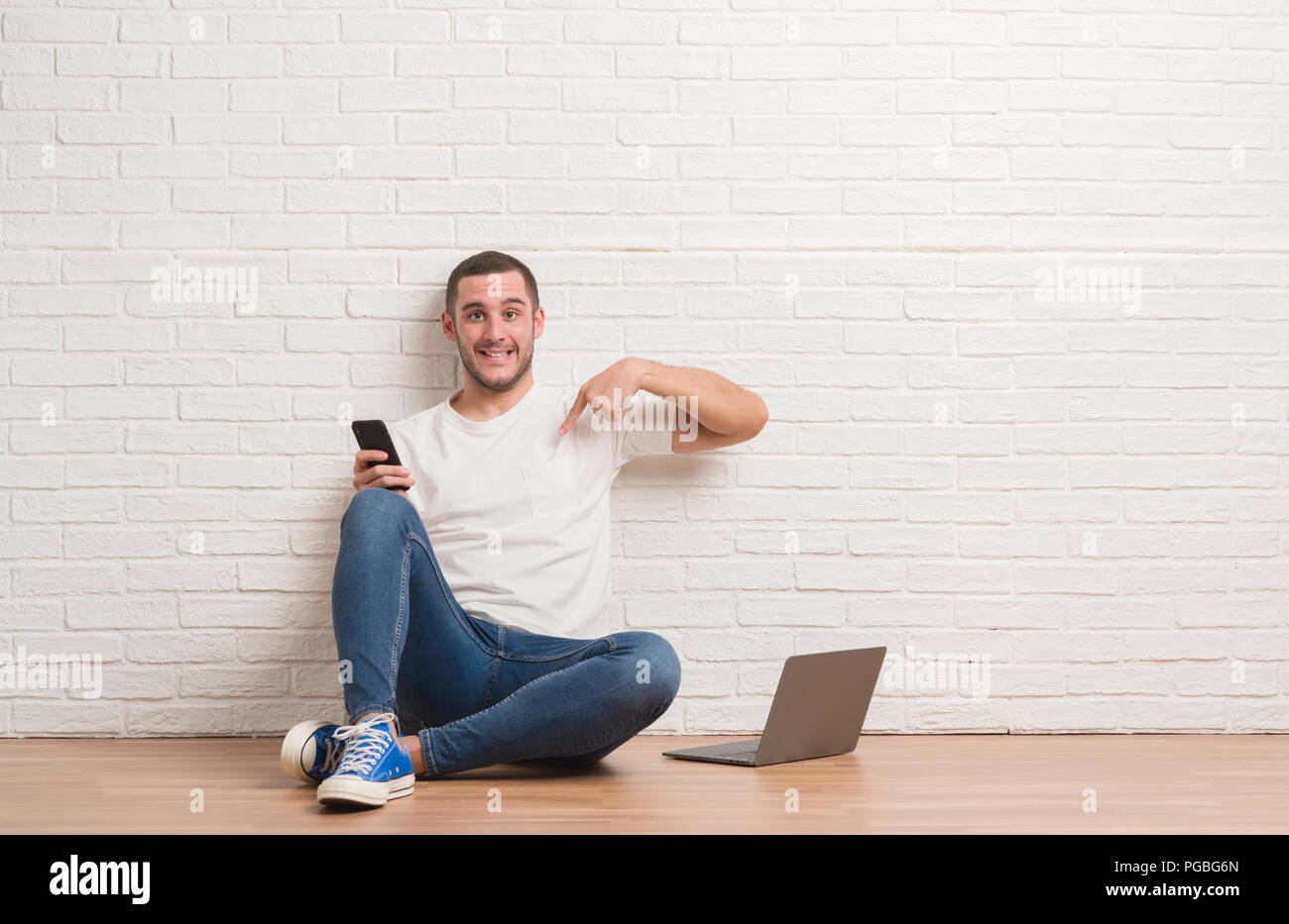 Young caucasian man sitting over white brick wall using computer laptop ...