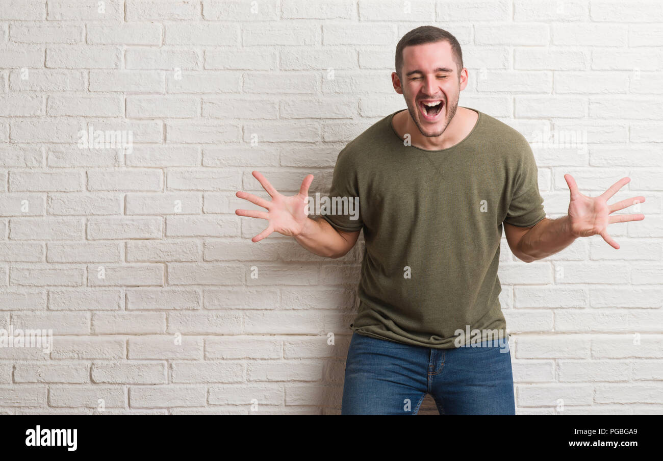 Young caucasian man standing over white brick wall celebrating mad and ...