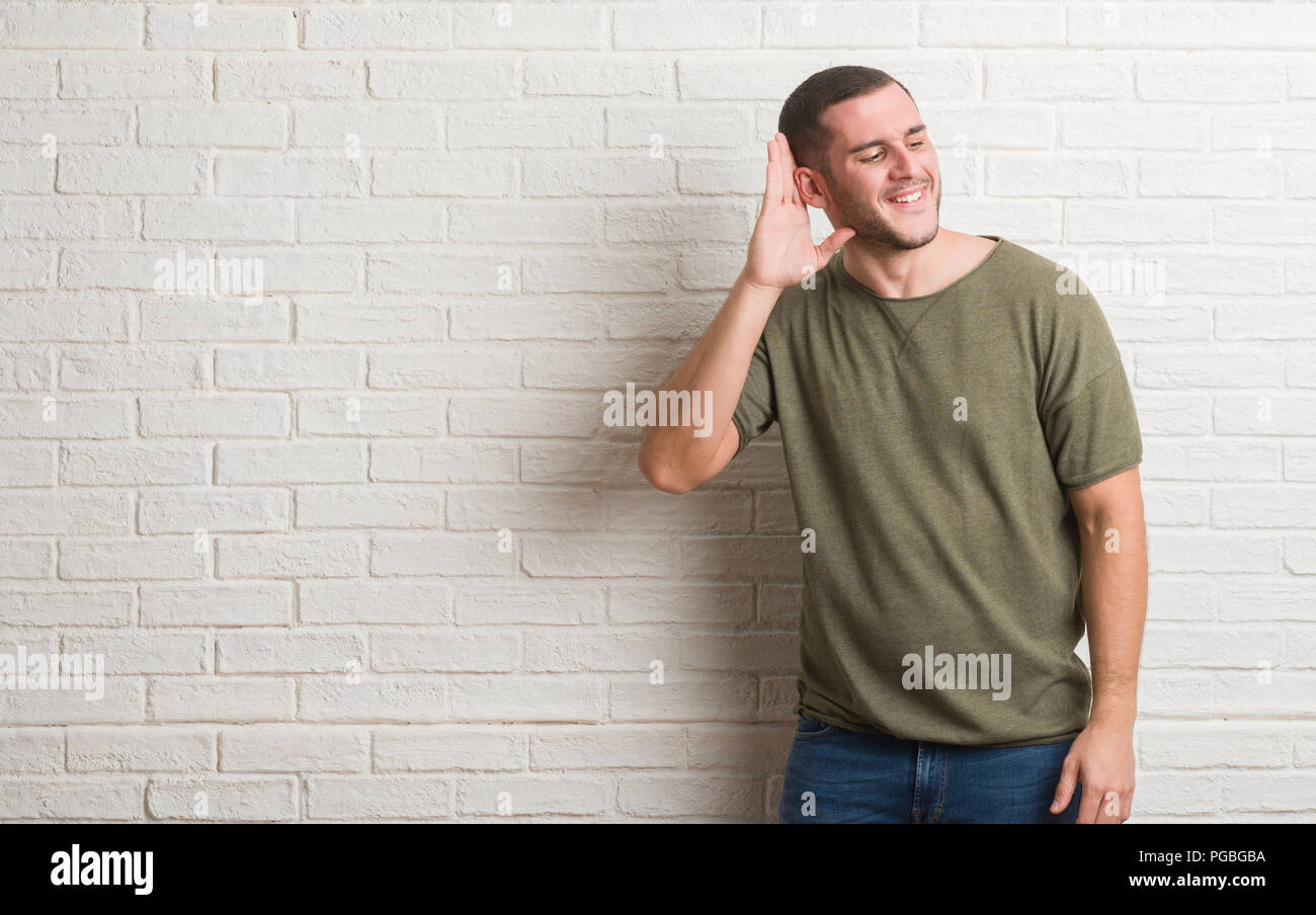 Young caucasian man standing over white brick wall smiling with hand ...