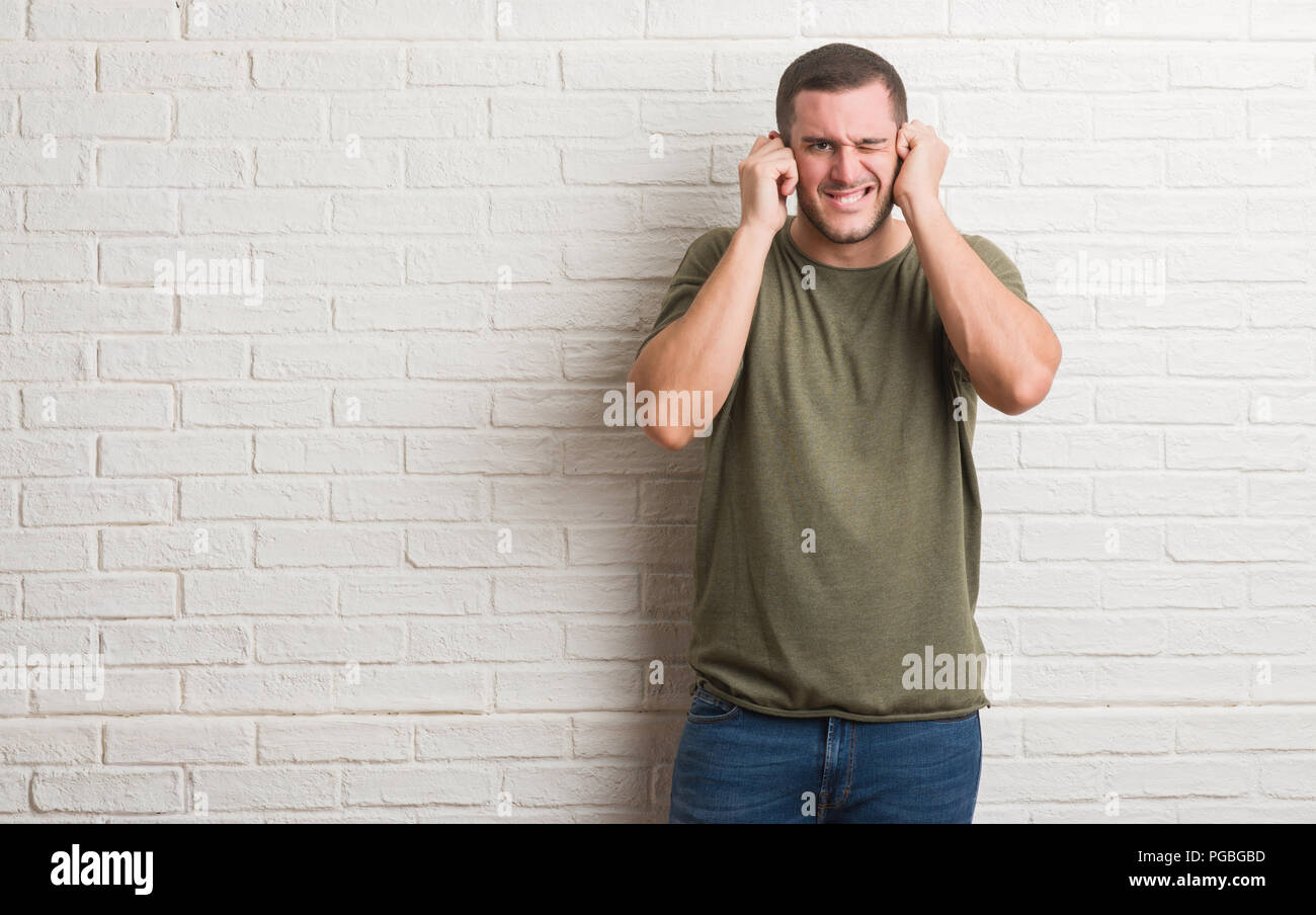 Young caucasian man standing over white brick wall covering ears with ...