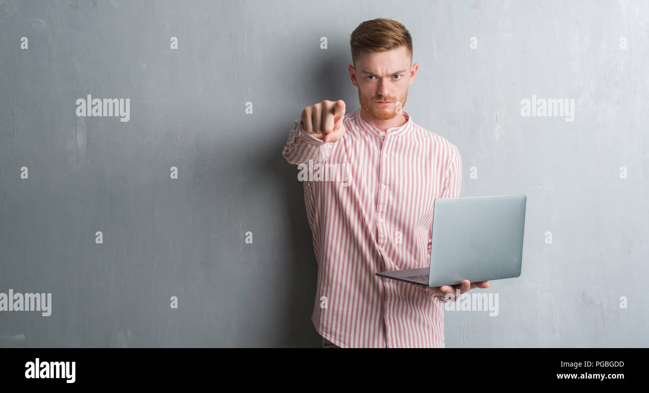 Young redhead man over grey grunge wall holding and using computer ...