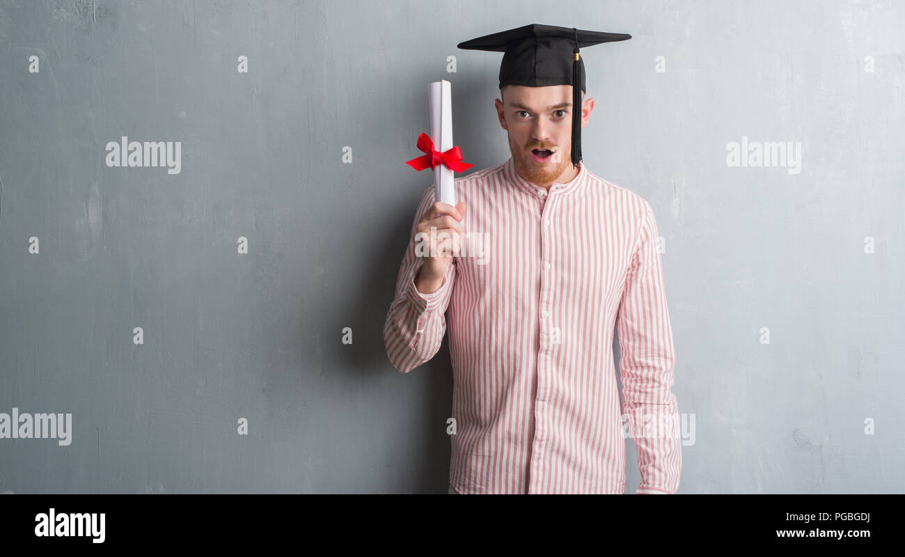 Young redhead man over grey grunge wall wearing graduate cap holding ...