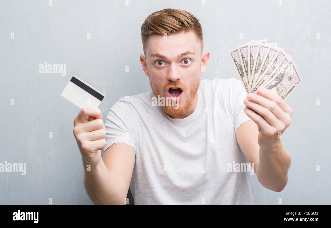 Young redhead man holding dollars and credit card scared in shock with ...